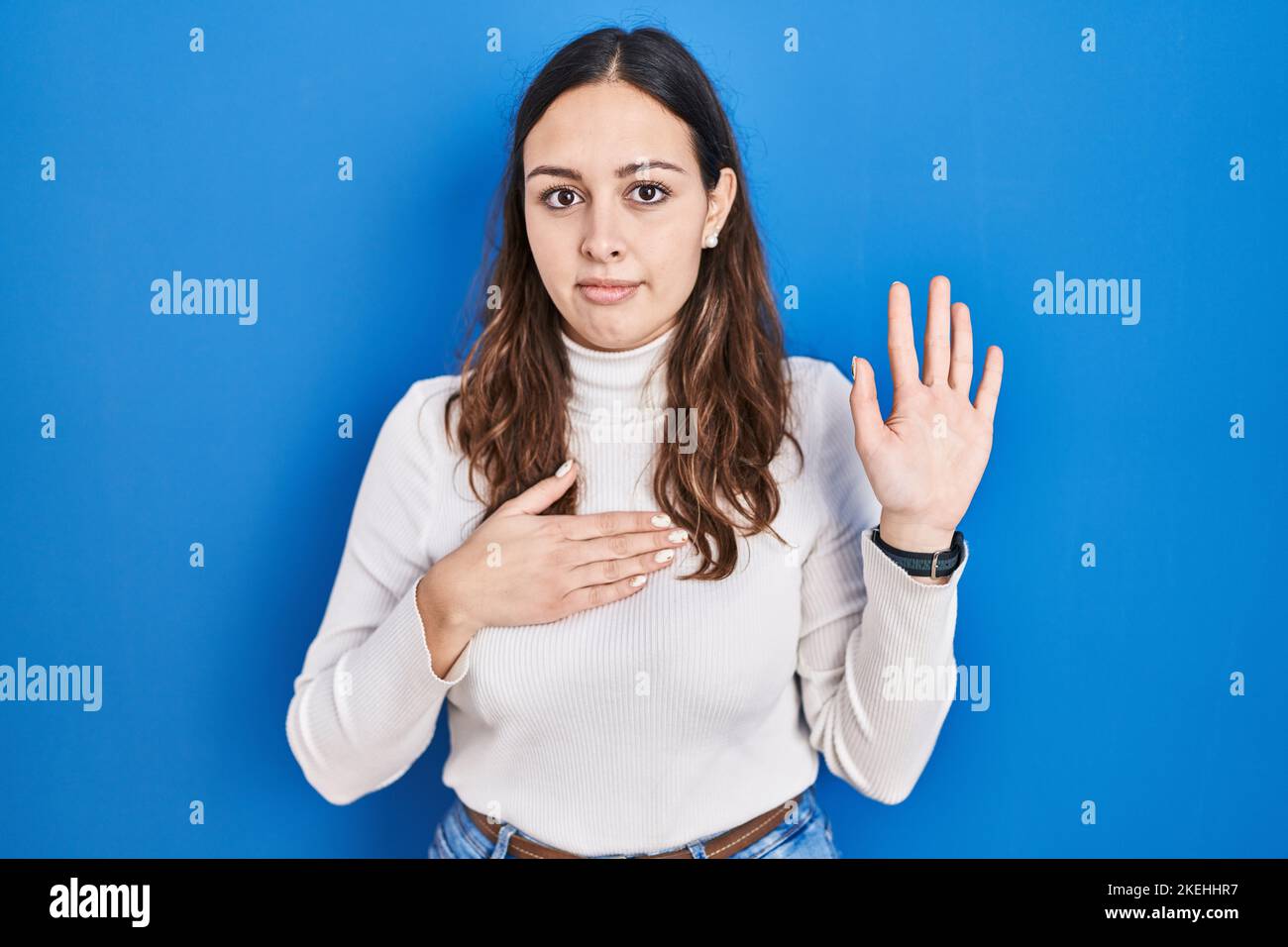 Young hispanic woman standing over blue background swearing with hand ...
