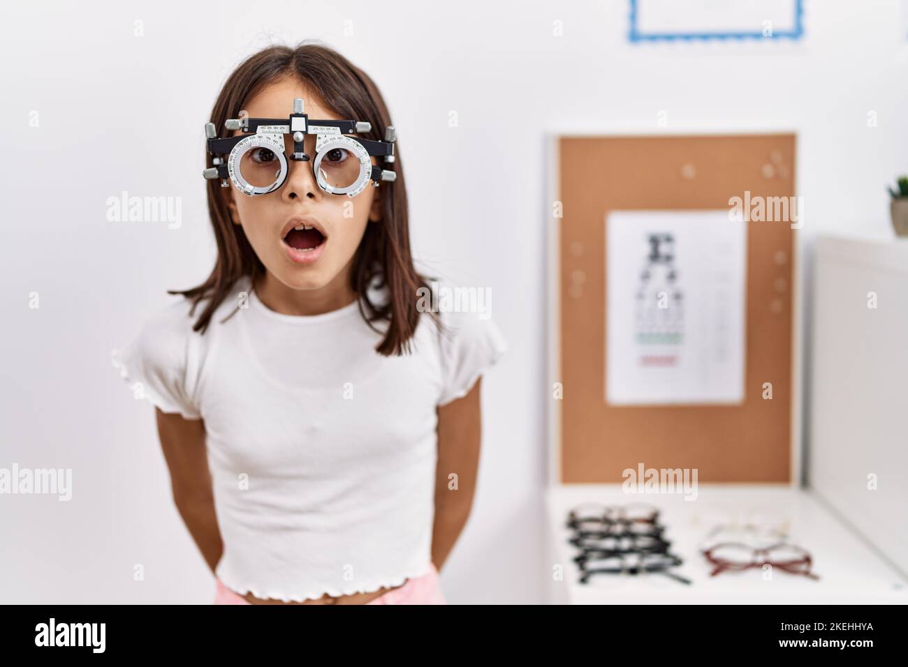Young hispanic girl wearing optometry glasses scared and amazed with ...