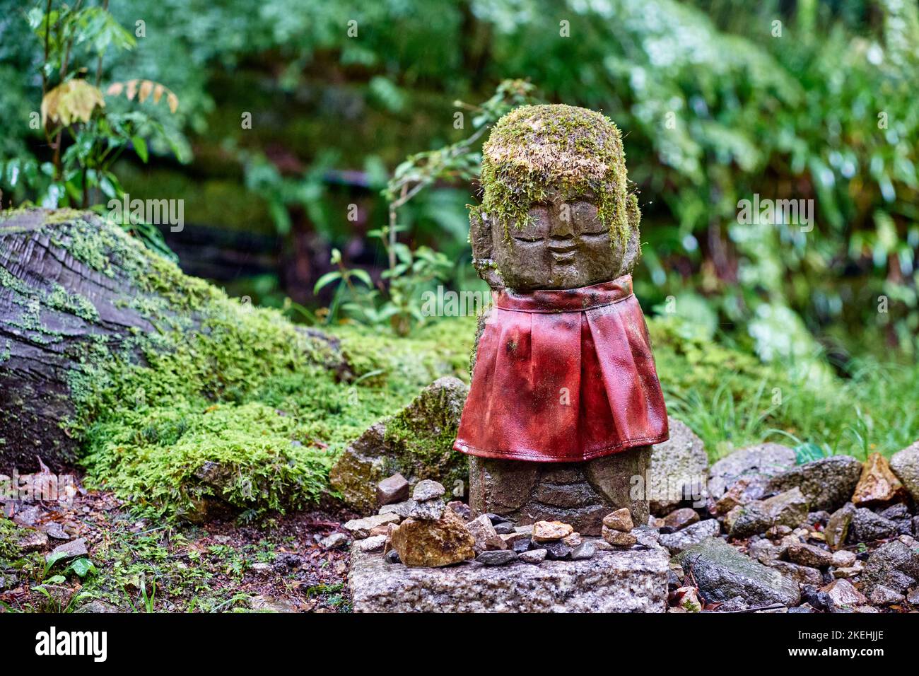 Moss covered jizo wearing red mantel standing eyes closed on a walk ...