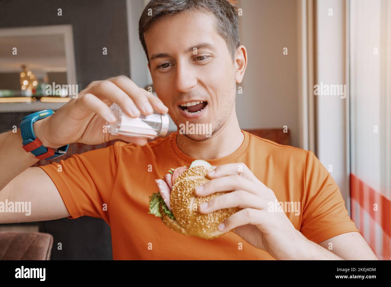 A man with a cheerful face pours an excess of salt on his burger in a ...