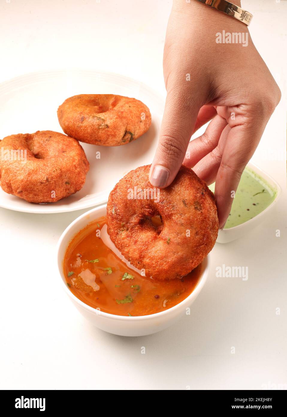 A vertical closeup of a hand dipping South Indian Medu vada in a sauce ...