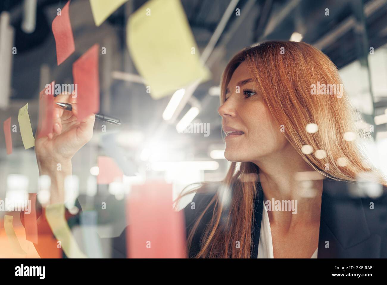 Businesswoman writing on sticky notes on glass wall while working in ...