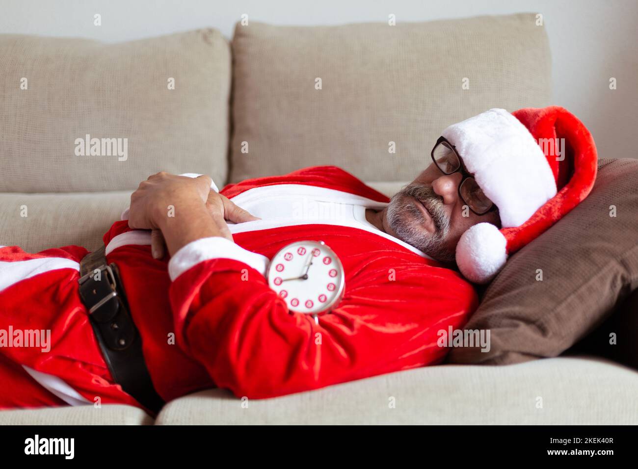 Older man dressed as Santa Claus sleeping on the sofa next to the alarm ...