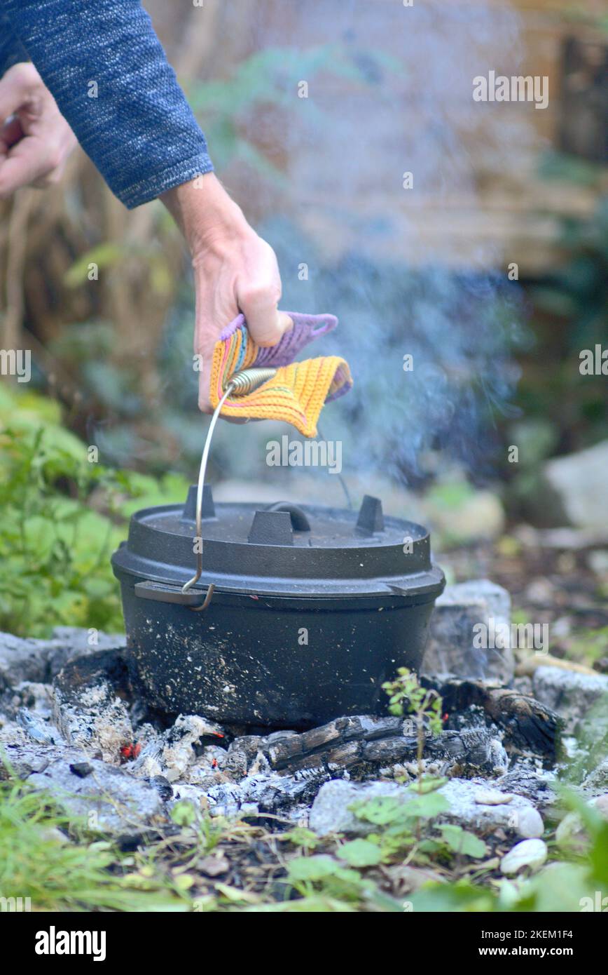A dutch pot on a charcoal camp fire with a man trying to remove the pot ...