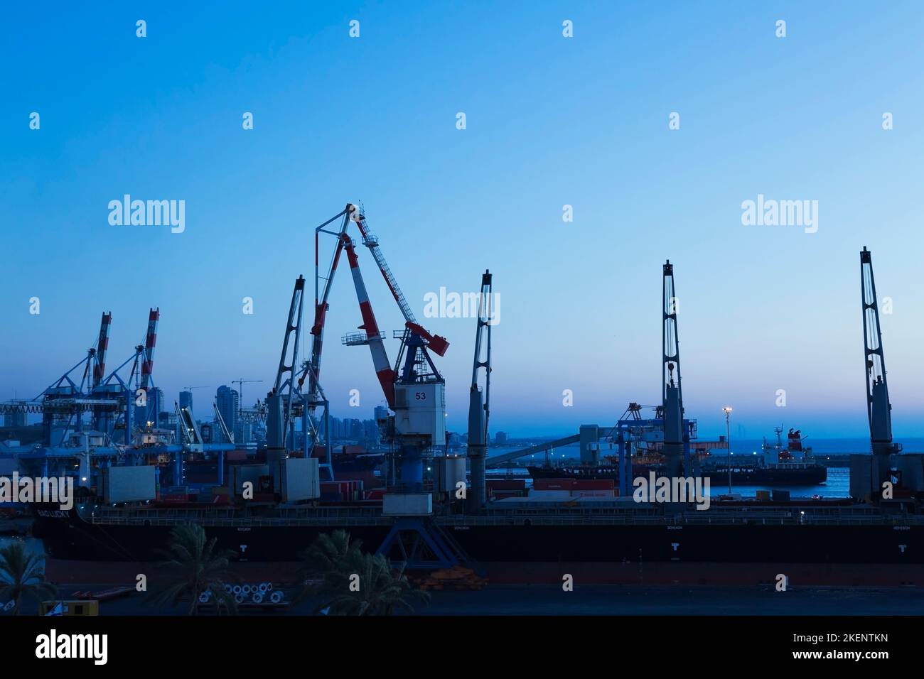 Silhouetted four-link cargo loading crane and docked cargo ship ...