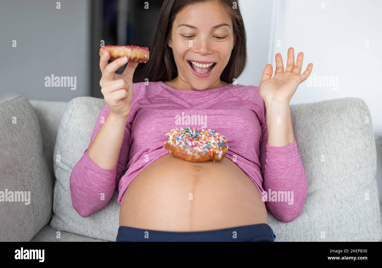 Pregnancy food cravings concept photo. Funny Asian pregnant woman eating donuts at home hungry ...