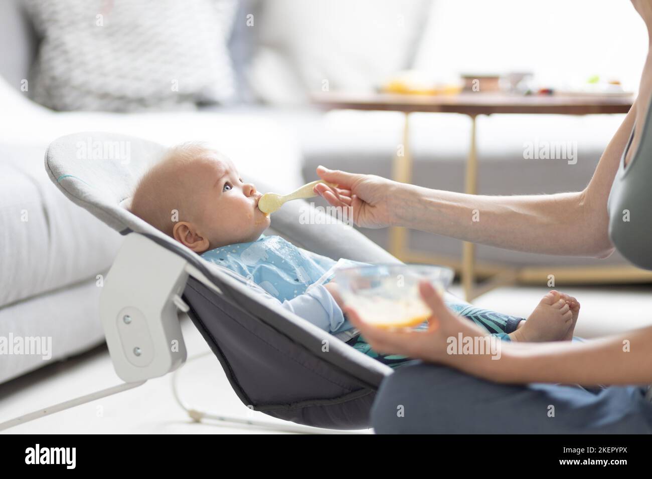 Mother spoon feeding her baby boy child in baby chair with fruit puree ...