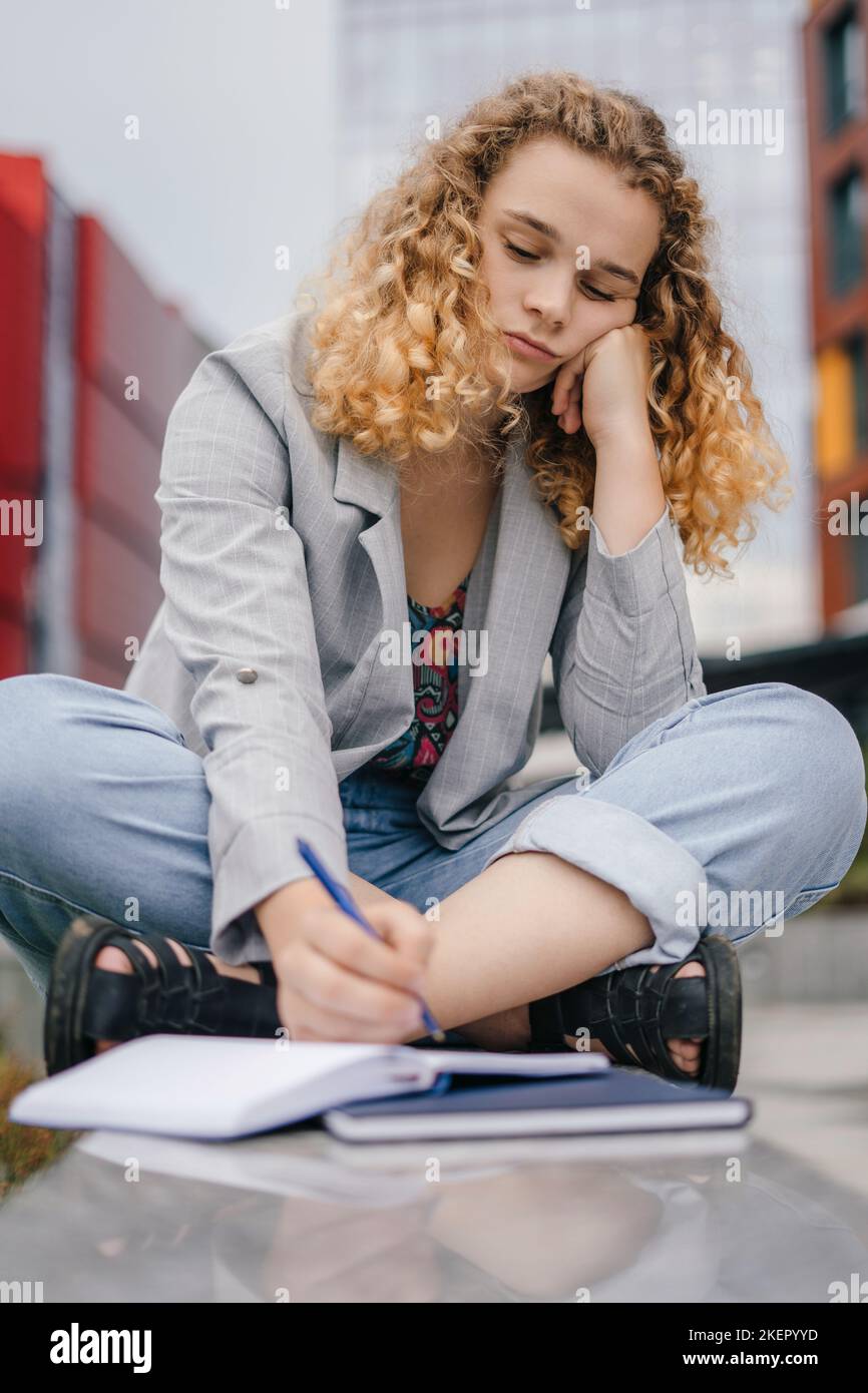Pensive woman with curly hair taking notes, writing resume on notepad ...