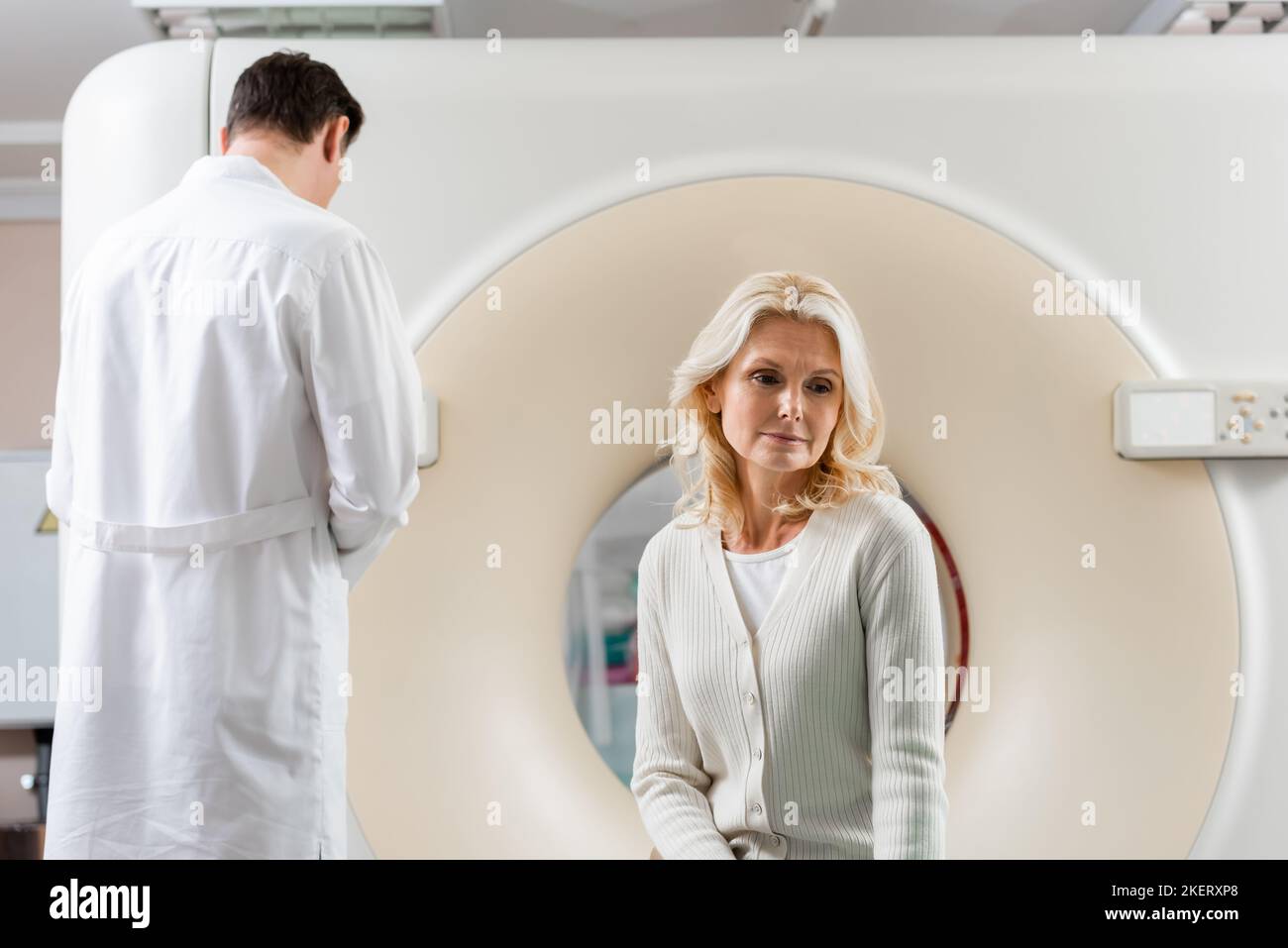 pensive blonde woman sitting near ct scanner and doctor in hospital ...