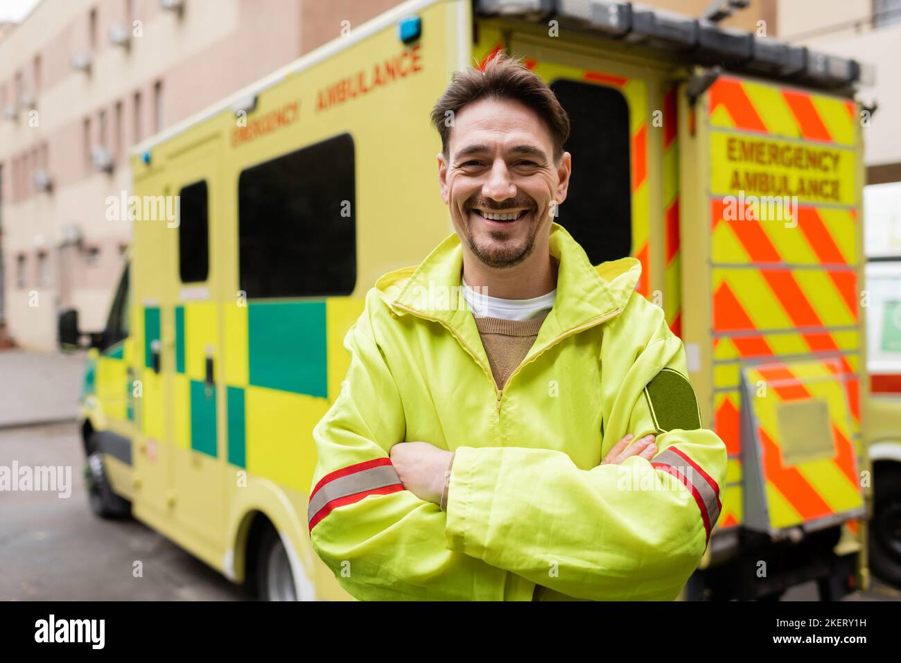 Smiling paramedic crossing arms and looking at camera near blurred ...