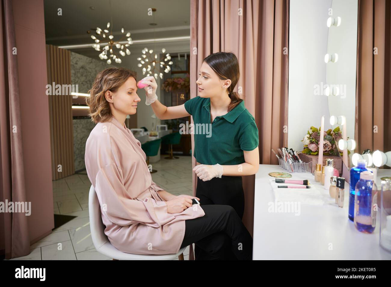 Young woman sitting at dressing table while makeup artist doing ...