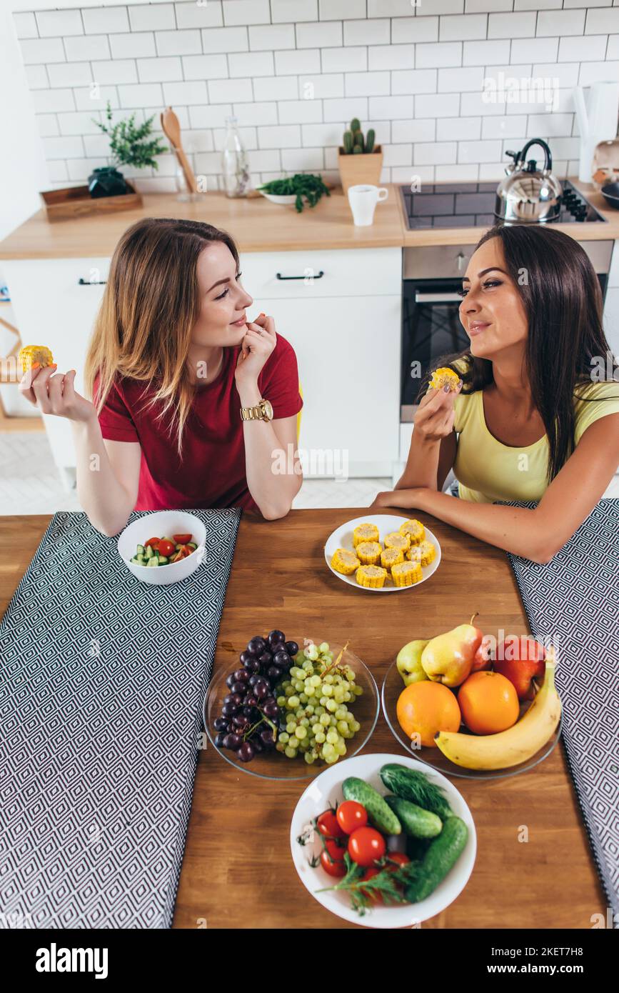 Two girls friends sitting in kitchen, eating fruits and vegetables ...