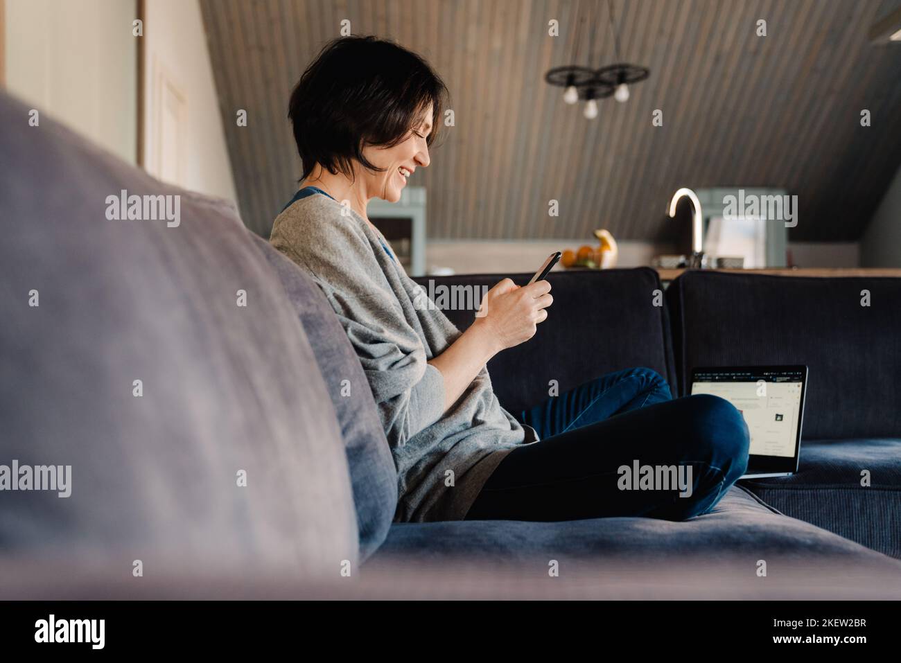 White mature woman using mobile phone while sitting on sofa at home ...