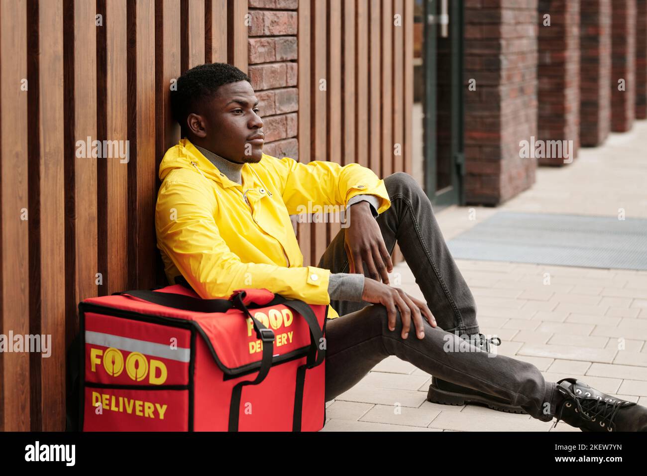 Young tired deliveryman in yellow jacket sitting on the road by brown ...