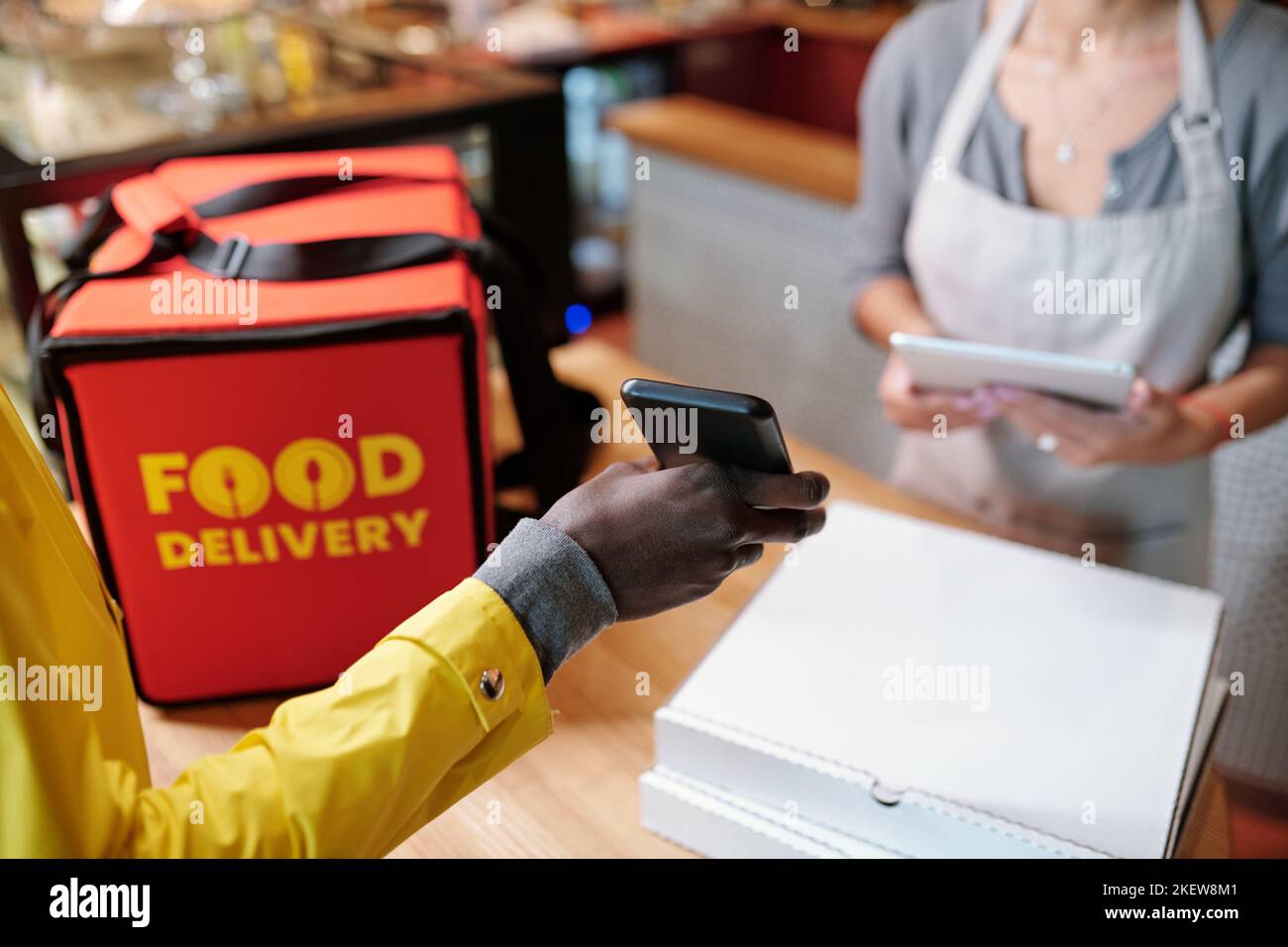 Hand of courier holding smartphone over table with big red bag and ...