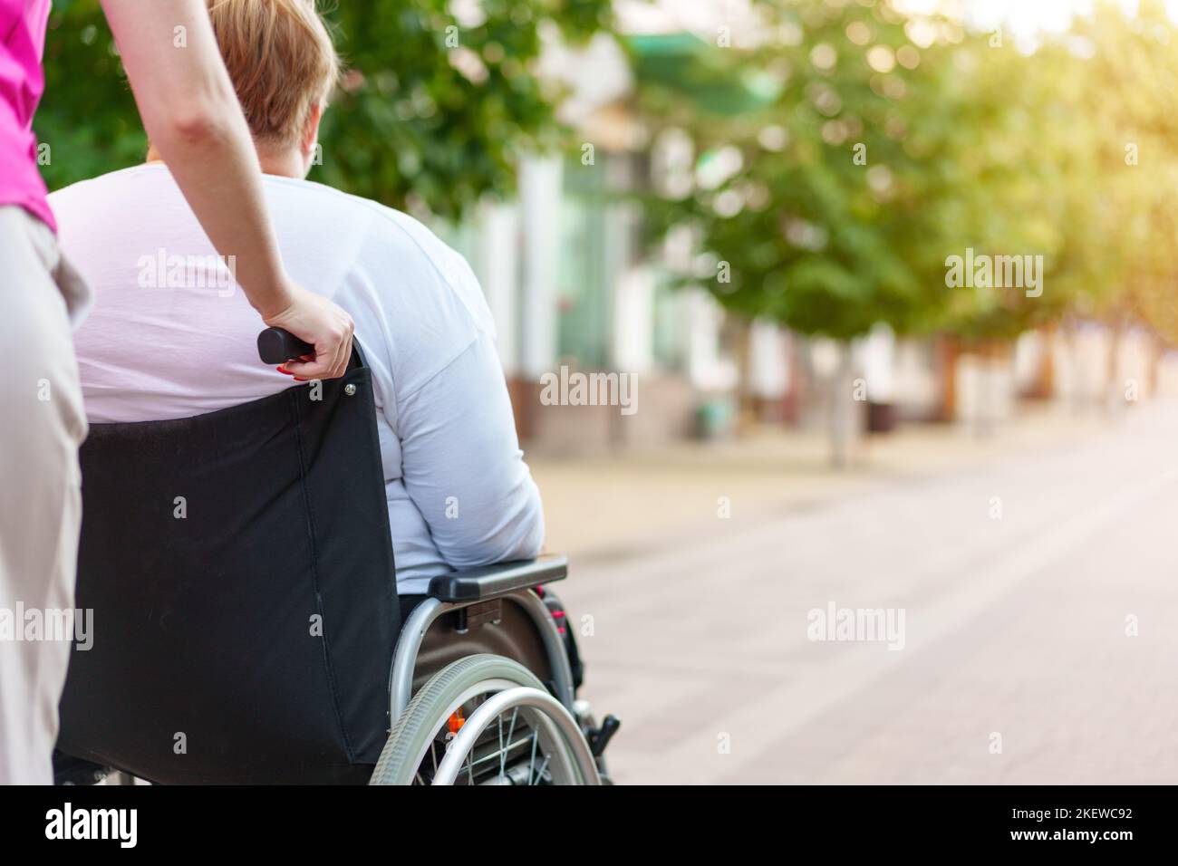 Back view of young woman helping mature woman in wheelchair in the city ...
