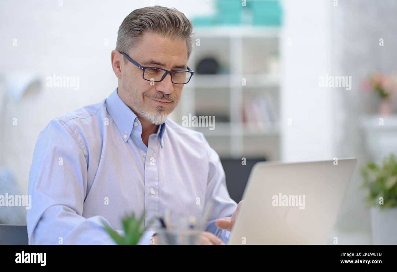 Older businessman sitting at desk in bright room working on laptop ...