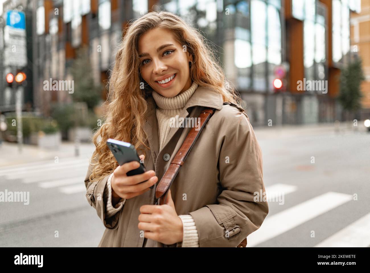 Smiling curly woman wearing warm coat walking down the street and using ...