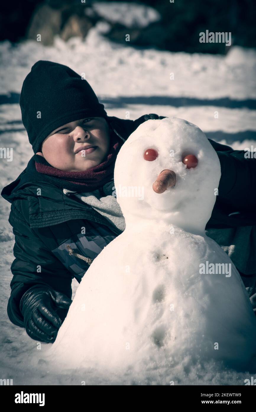 boy lying in the snow next to self-made snowman with carrot nose and ...