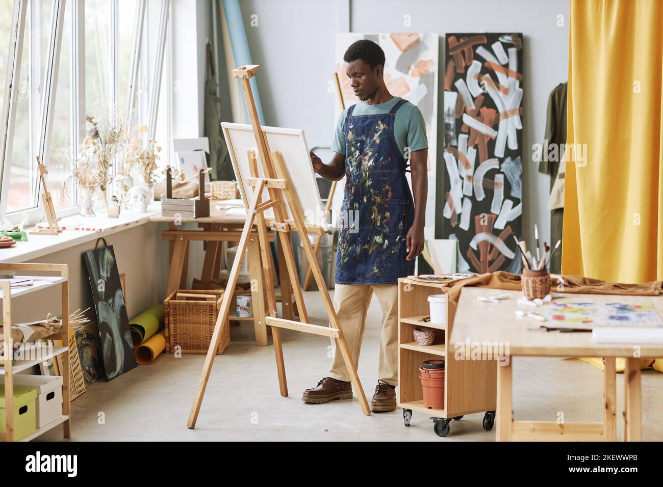 Young serious creative male artist in apron standing in front of easel ...