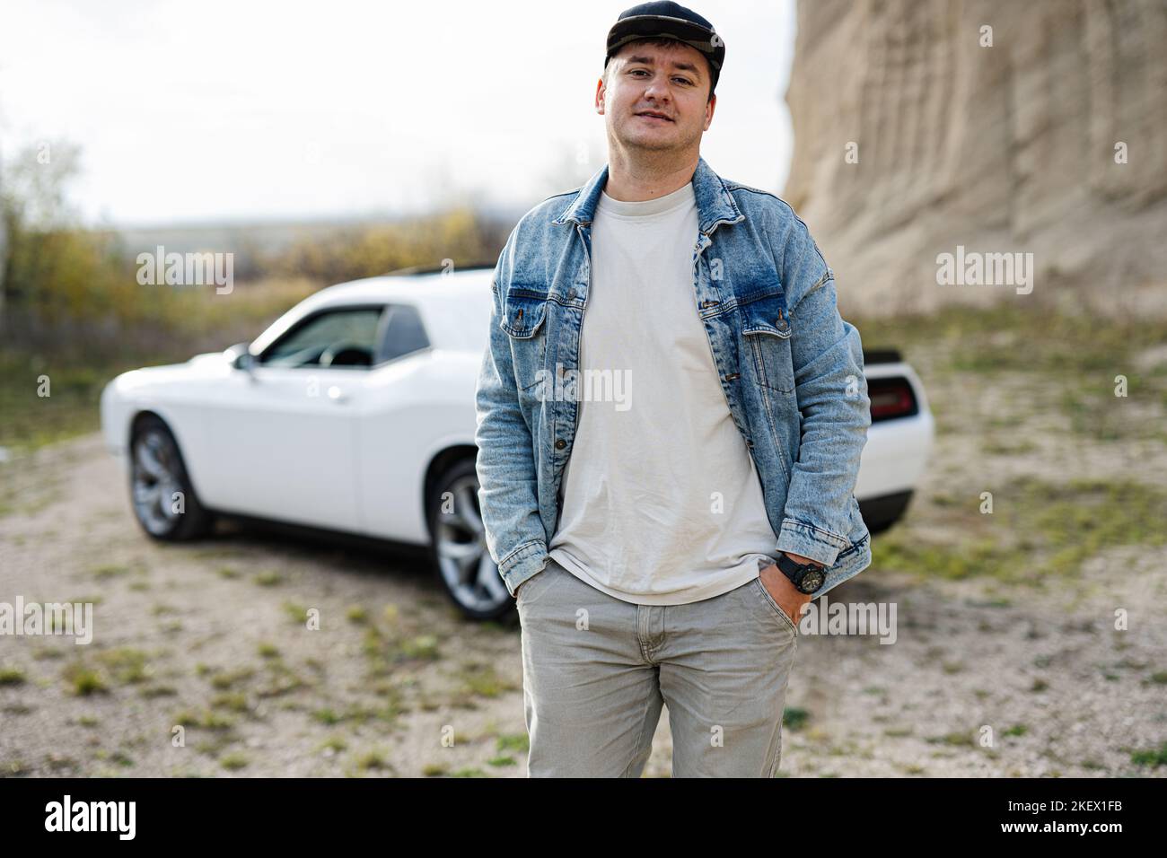 Handsome man in jeans jacket and cap walking near his white muscle car ...