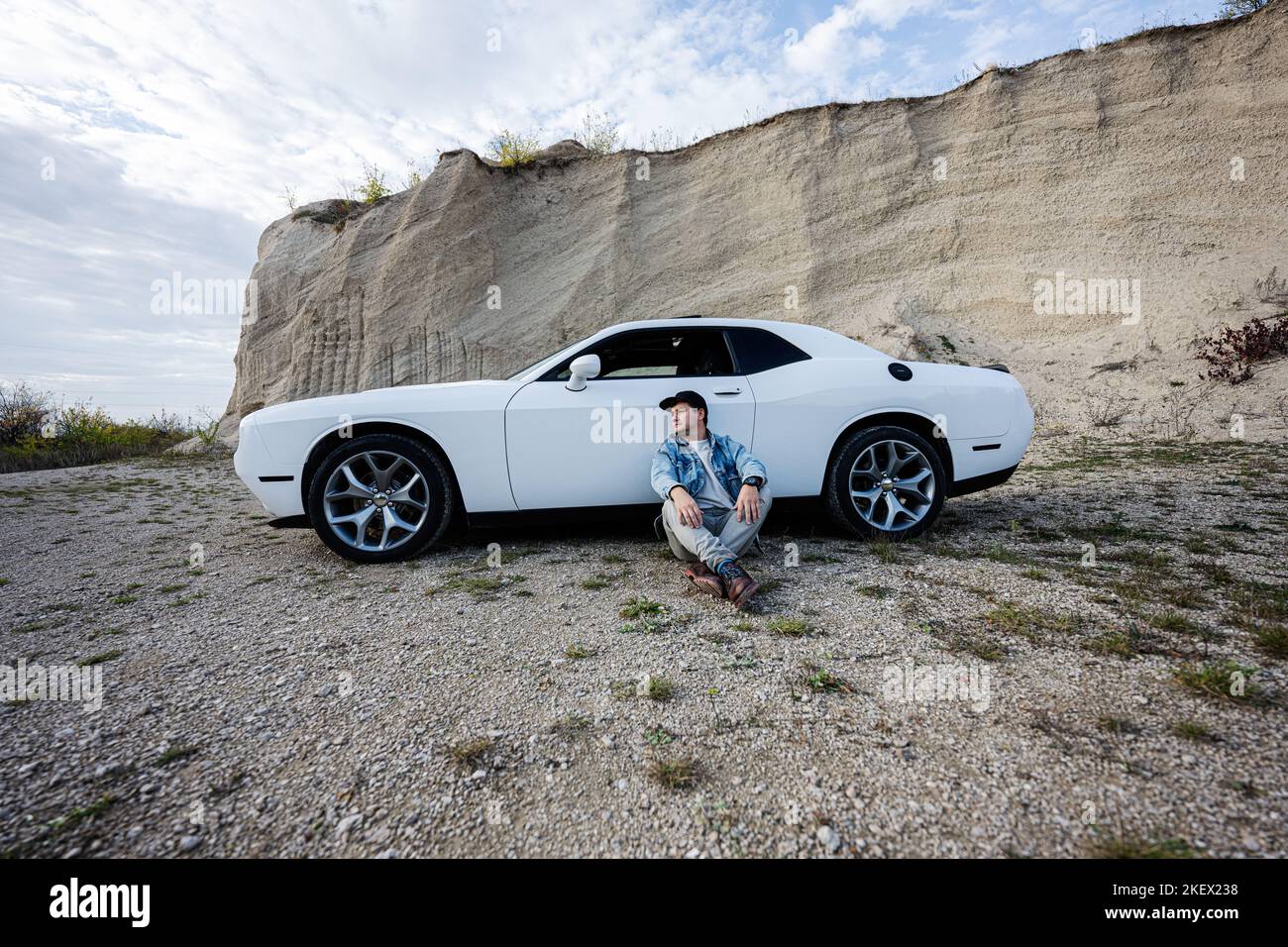 Handsome man in jeans jacket and cap sitting near his white muscle car ...