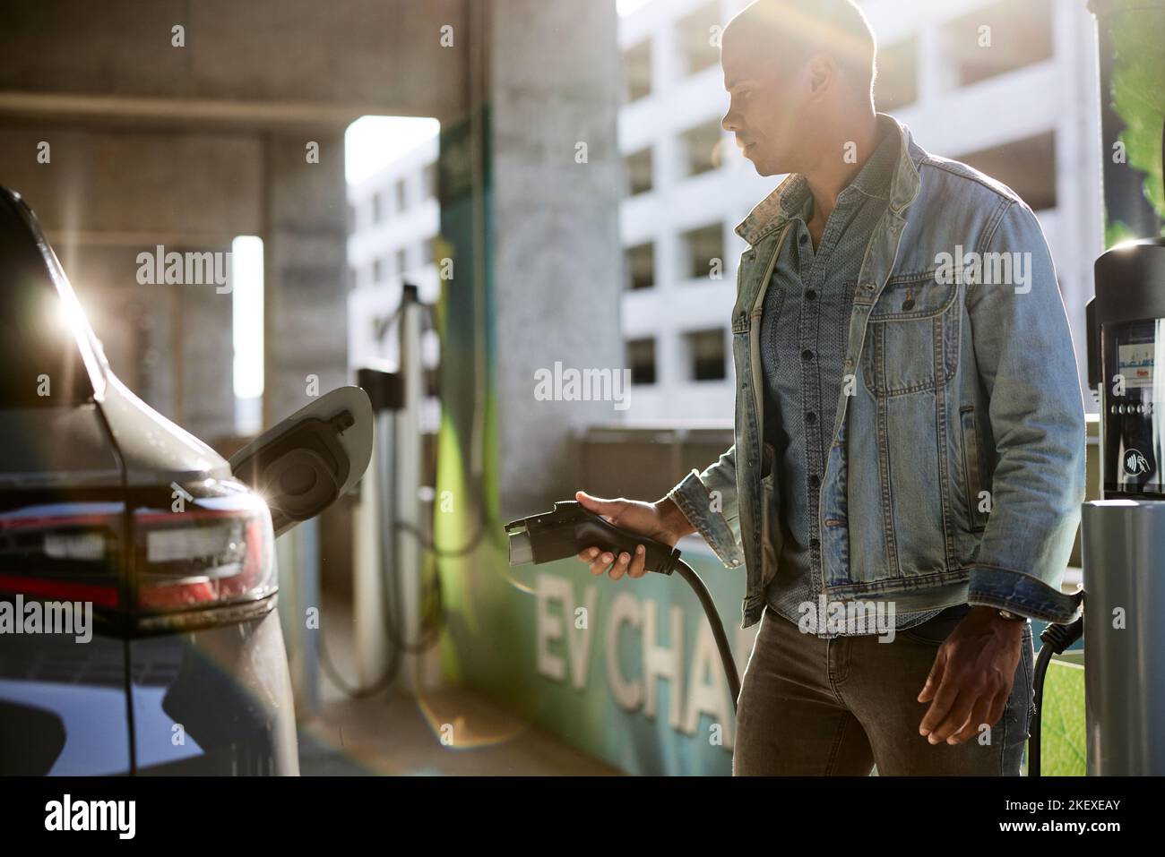 Man in denim jacket holding electric plug by car at charging station ...