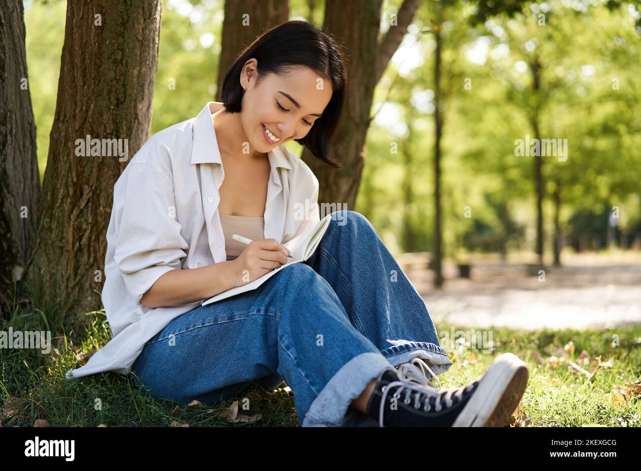 Portrait of happy asian girl sitting in park and writing in her diary ...