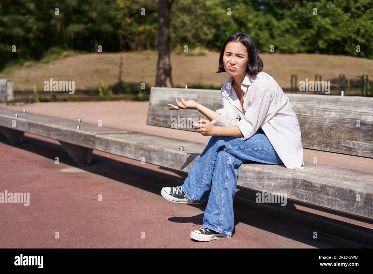Depressed asian girl sits on bench in park with smartphone, feeling ...