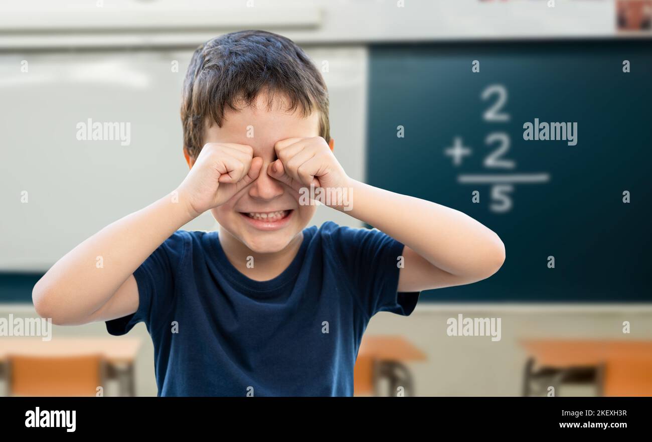 Little boy in blue t-shirt in class room with sad expression covering ...