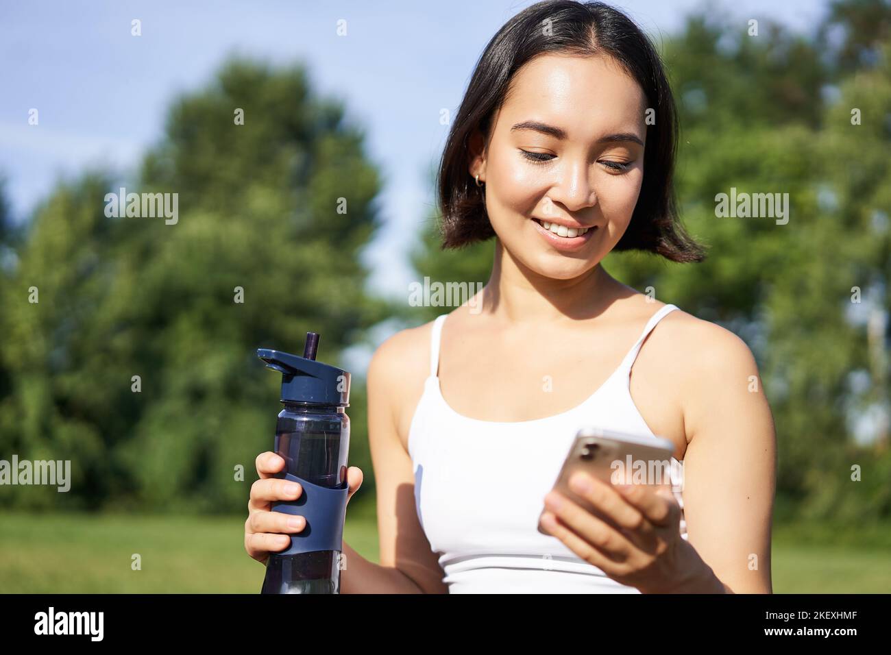 Smiling fitness girl drinks water, checks her app on smartphone and ...