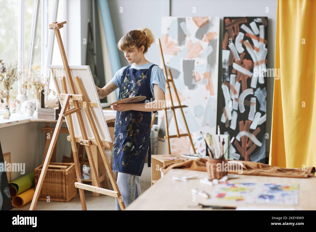 Young blond craftswoman in apron painting new masterpiece on canvas in ...
