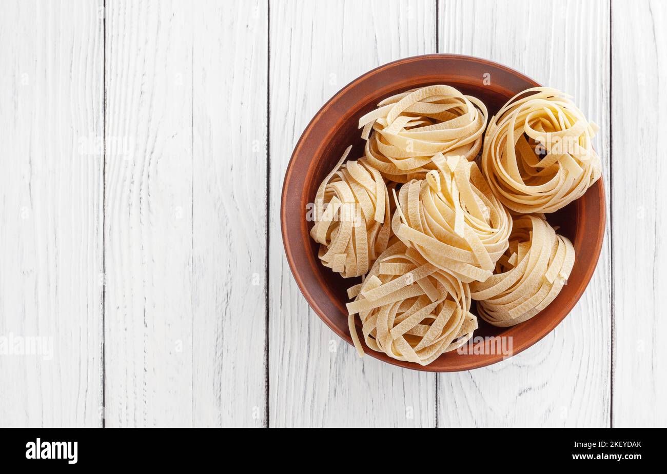 Uncooked tagliatelle pasta in ceramic bowl on white wooden background ...