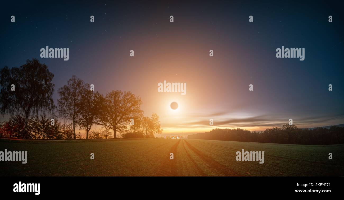 total solar eclipse during sunrise over a green field and trees along ...