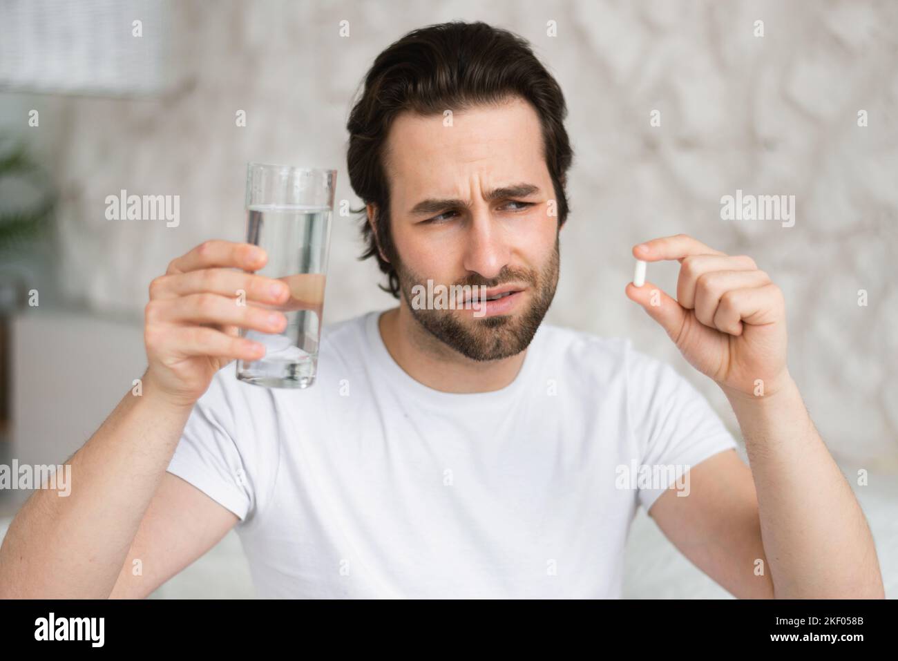 Portrait of sad millennial caucasian man with stubble sits on bed holds ...