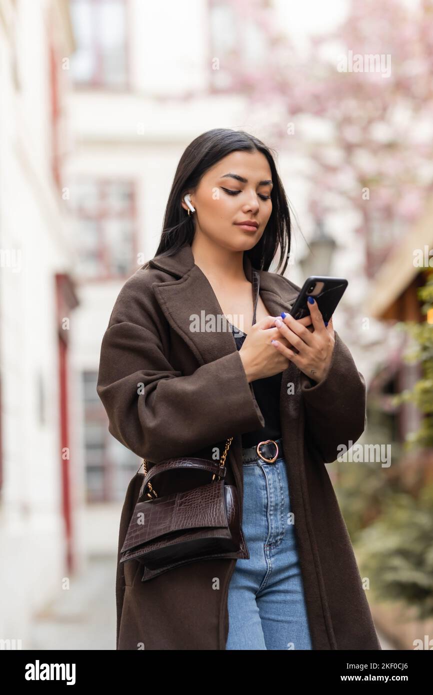 trendy brunette woman in wireless earphone using cellphone on street in ...