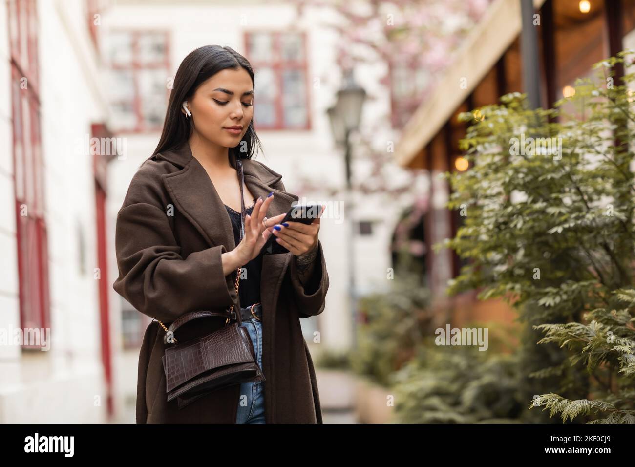 brunette woman in coat and wireless earphone using smartphone near ...