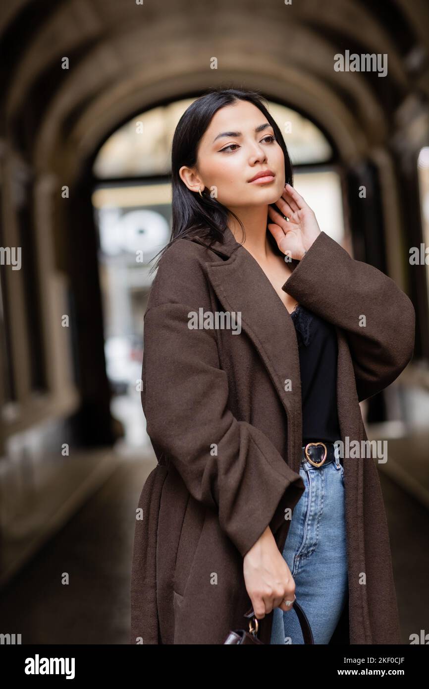 brunette woman in brown coat touching neck and looking away on blurred ...