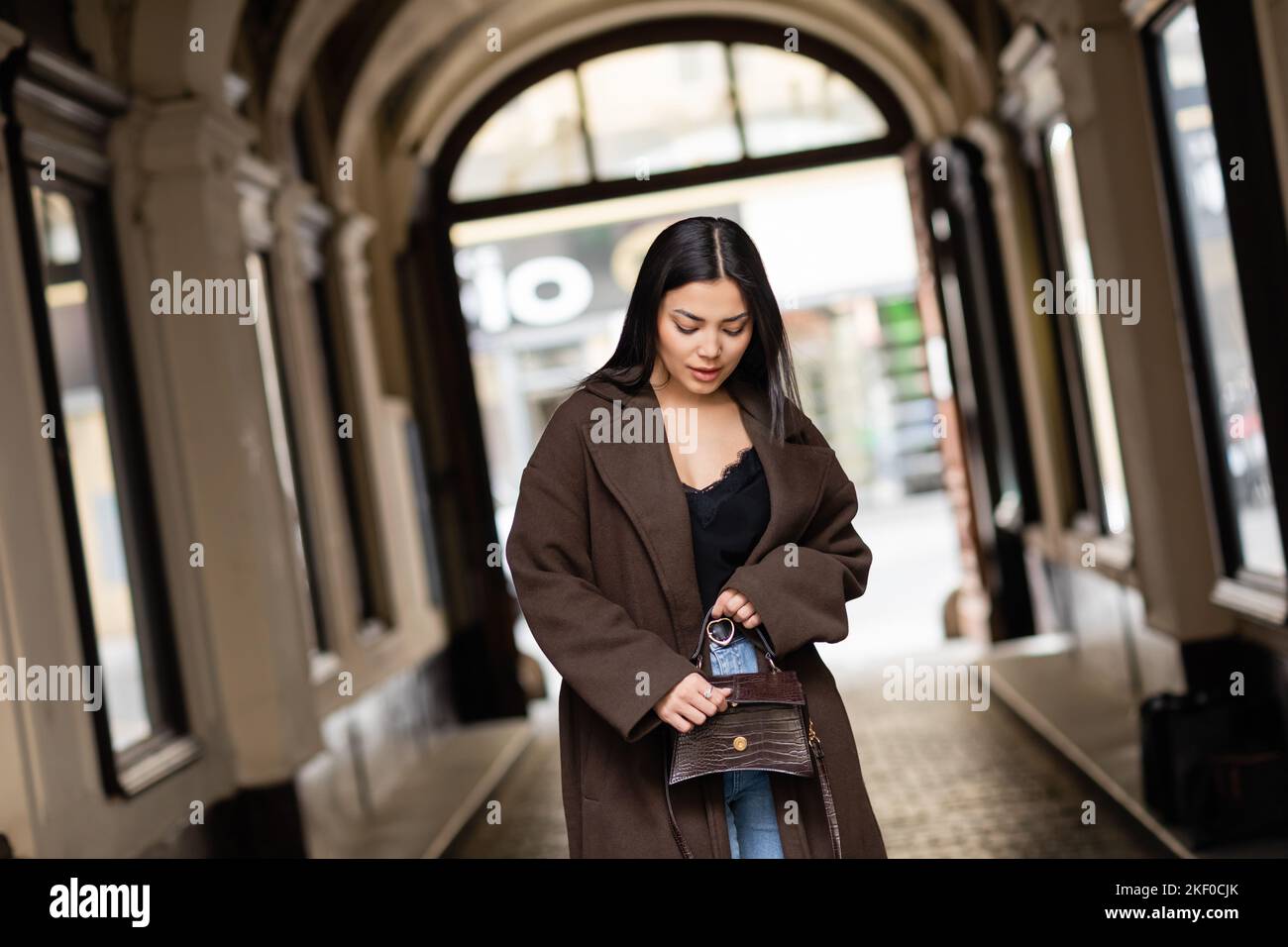 young woman in autumn coat opening handbag near arch building on ...