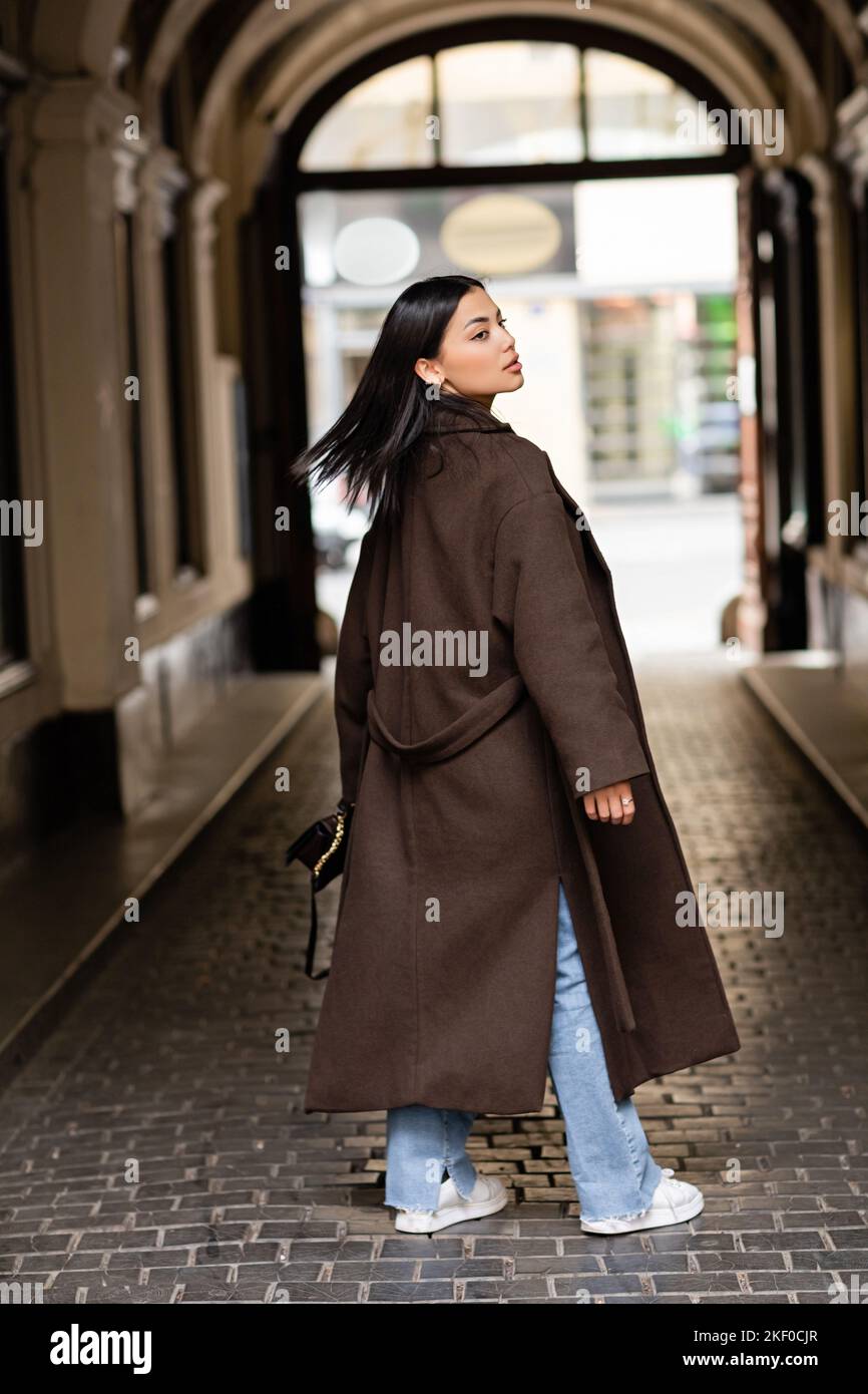 back view of young brunette woman in brown coat looking away under arch ...