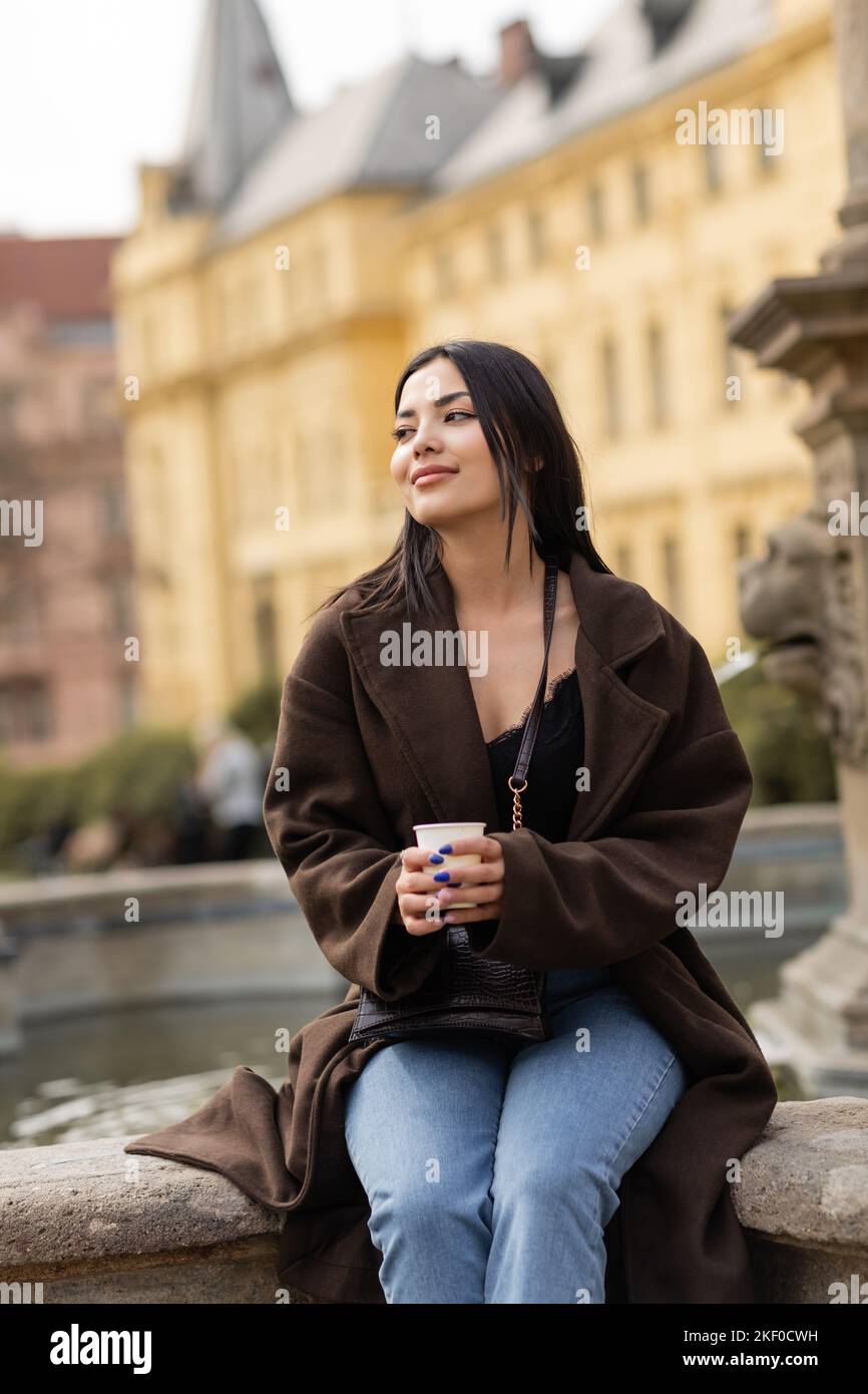 Positive brunette woman in coat holding paper cup near blurred fountain ...