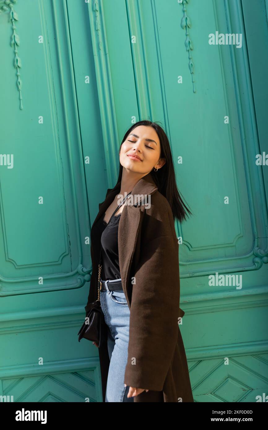 Pleased woman in coat standing in sunlight near turquoise door of ...