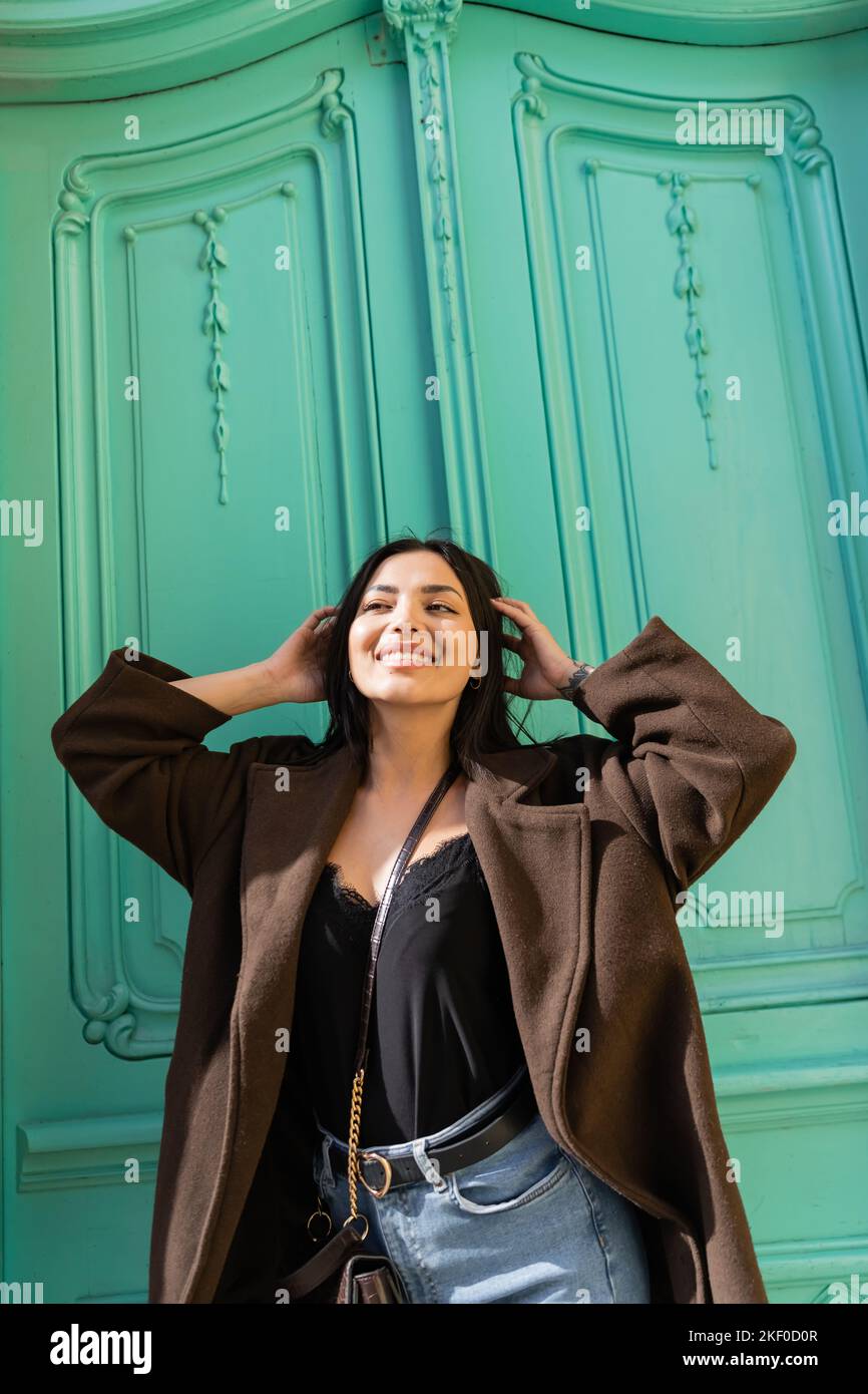 Happy young woman in coat touching hair near turquoise door of building ...
