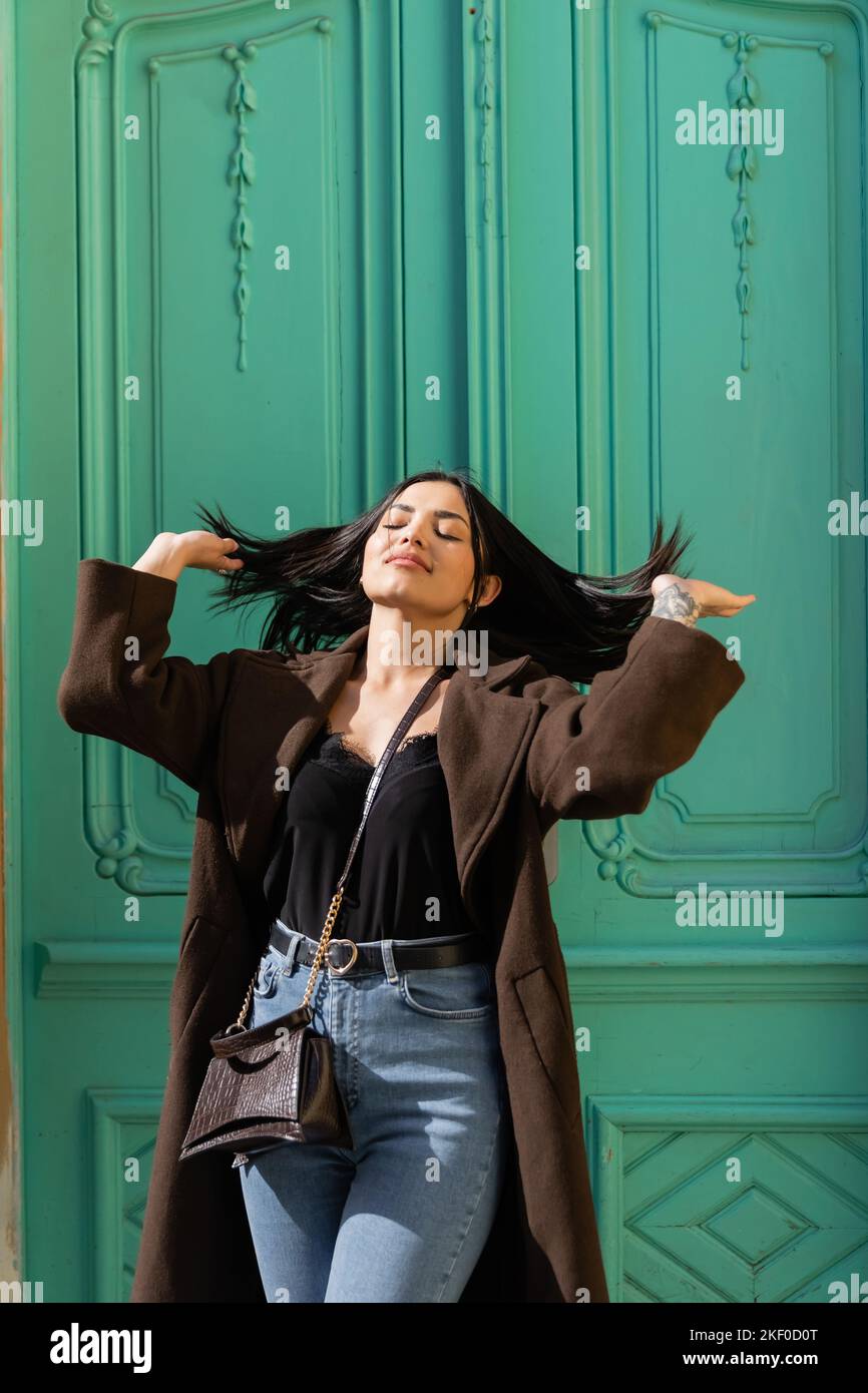 Young woman in coat touching hair near turquoise door of building on ...