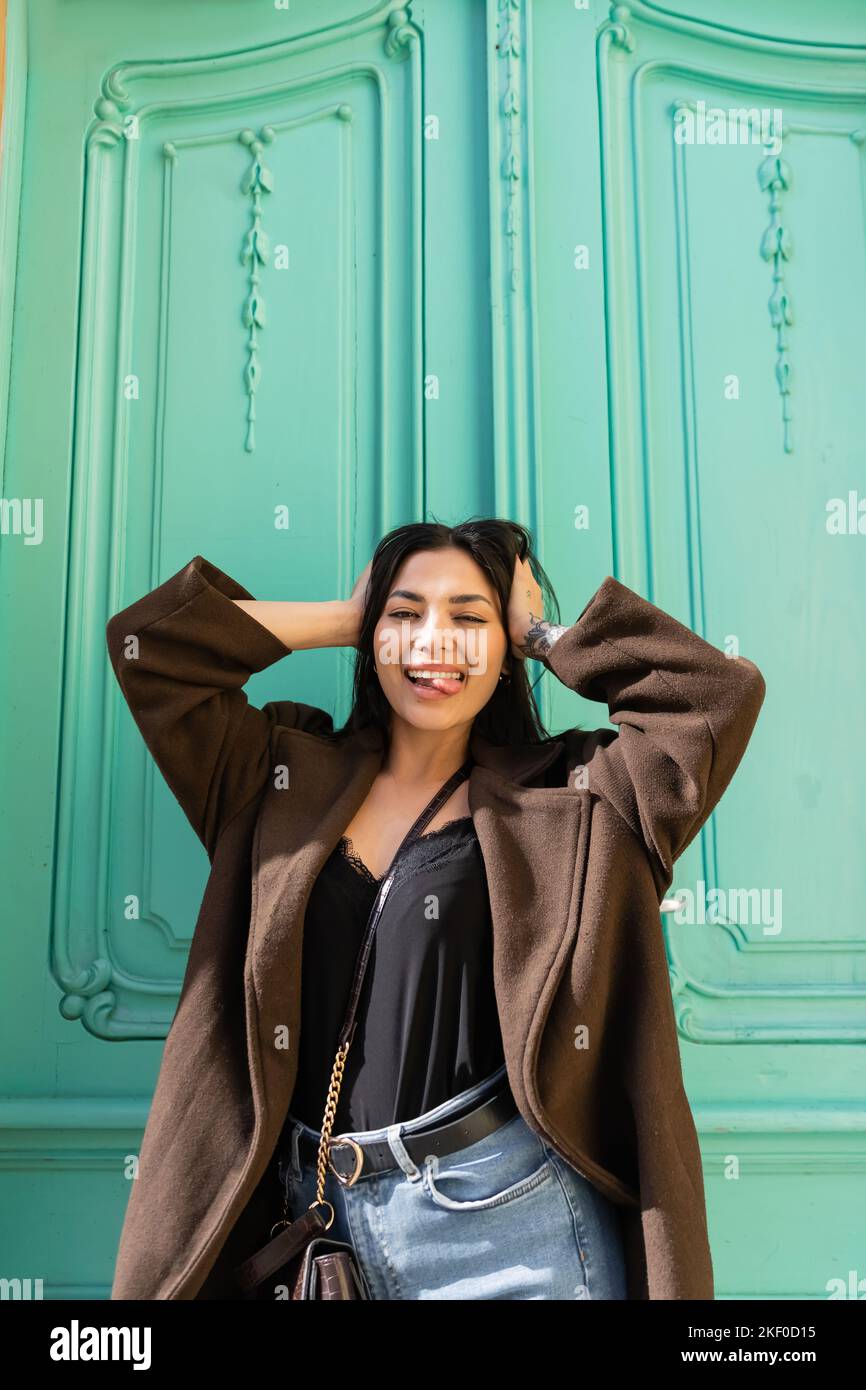 Young woman in coat sticking out tongue at camera near turquoise door ...