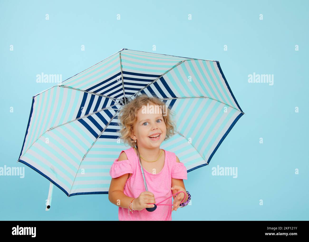Cheerful little girl stand under open umbrella on empty blue background ...