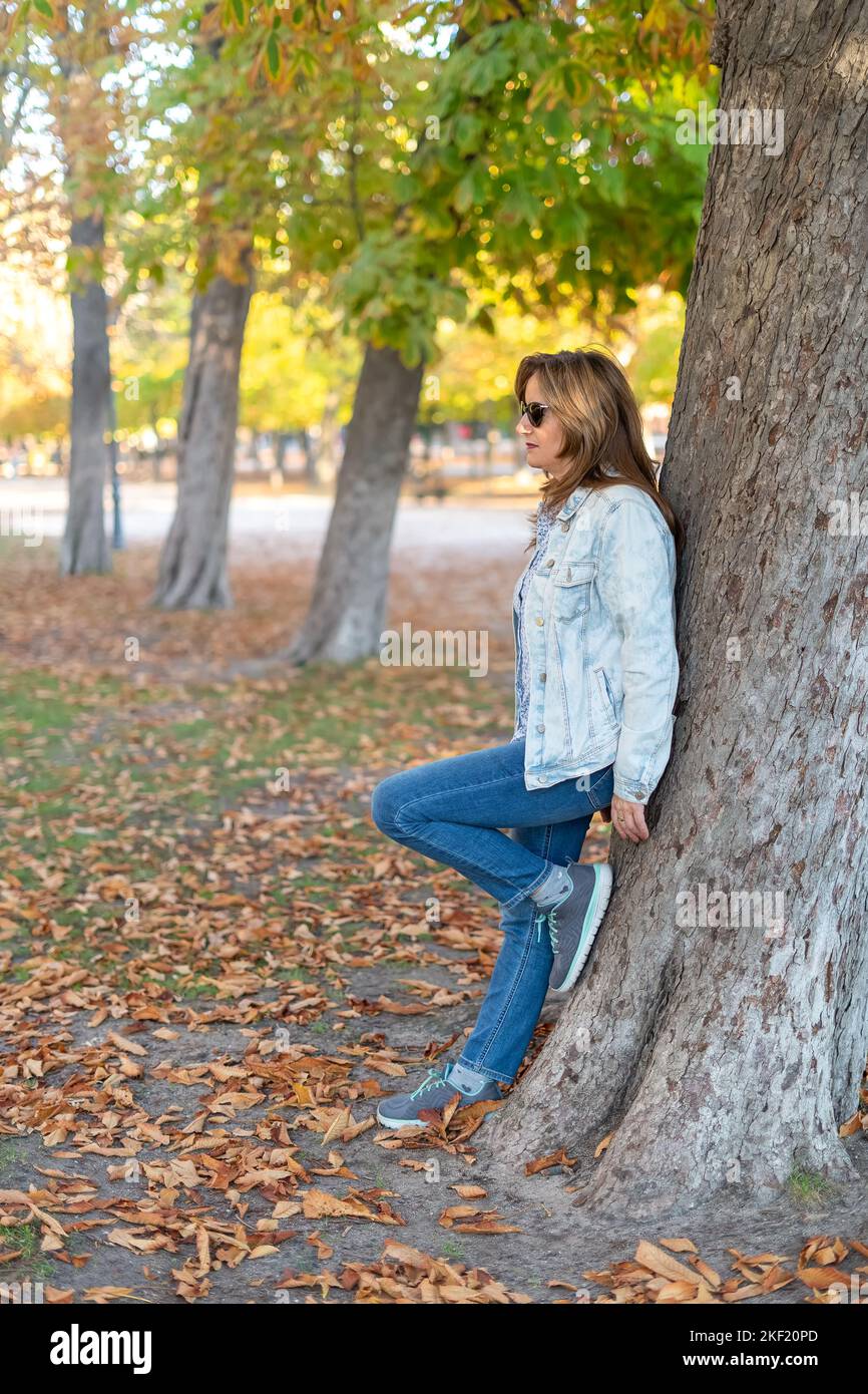 Mature white woman leaning against a large tree in a public park with ...
