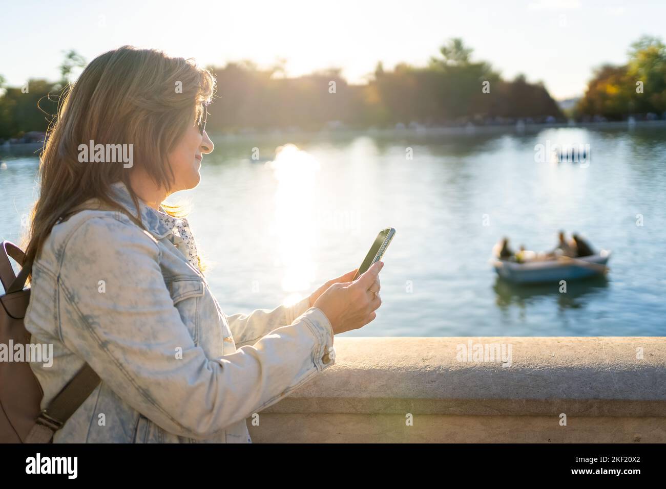 Woman looking at her mobile phone next to a lake in the park and with ...