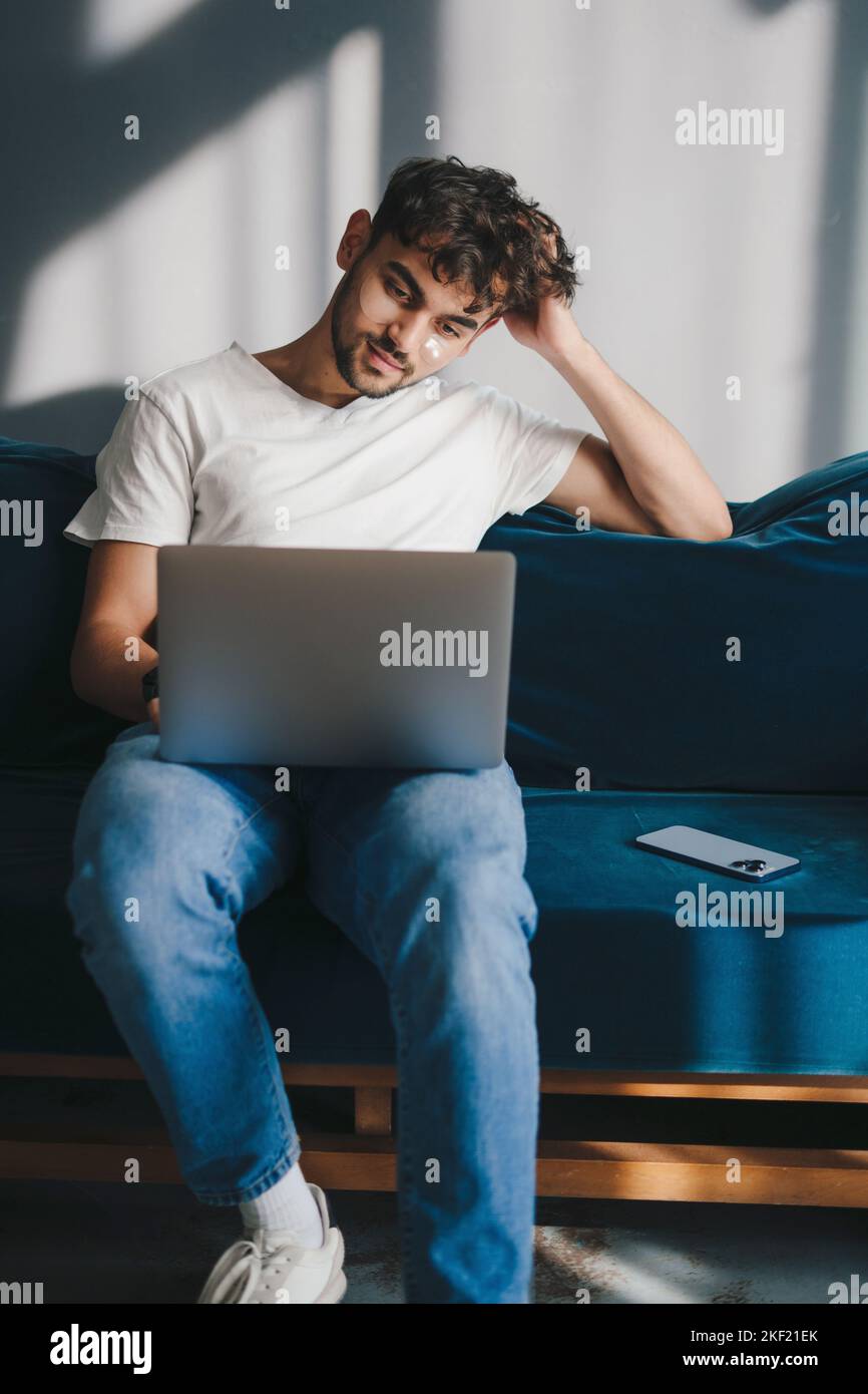 Relaxed young man working on laptop pc computer sitting on blue sofa ...