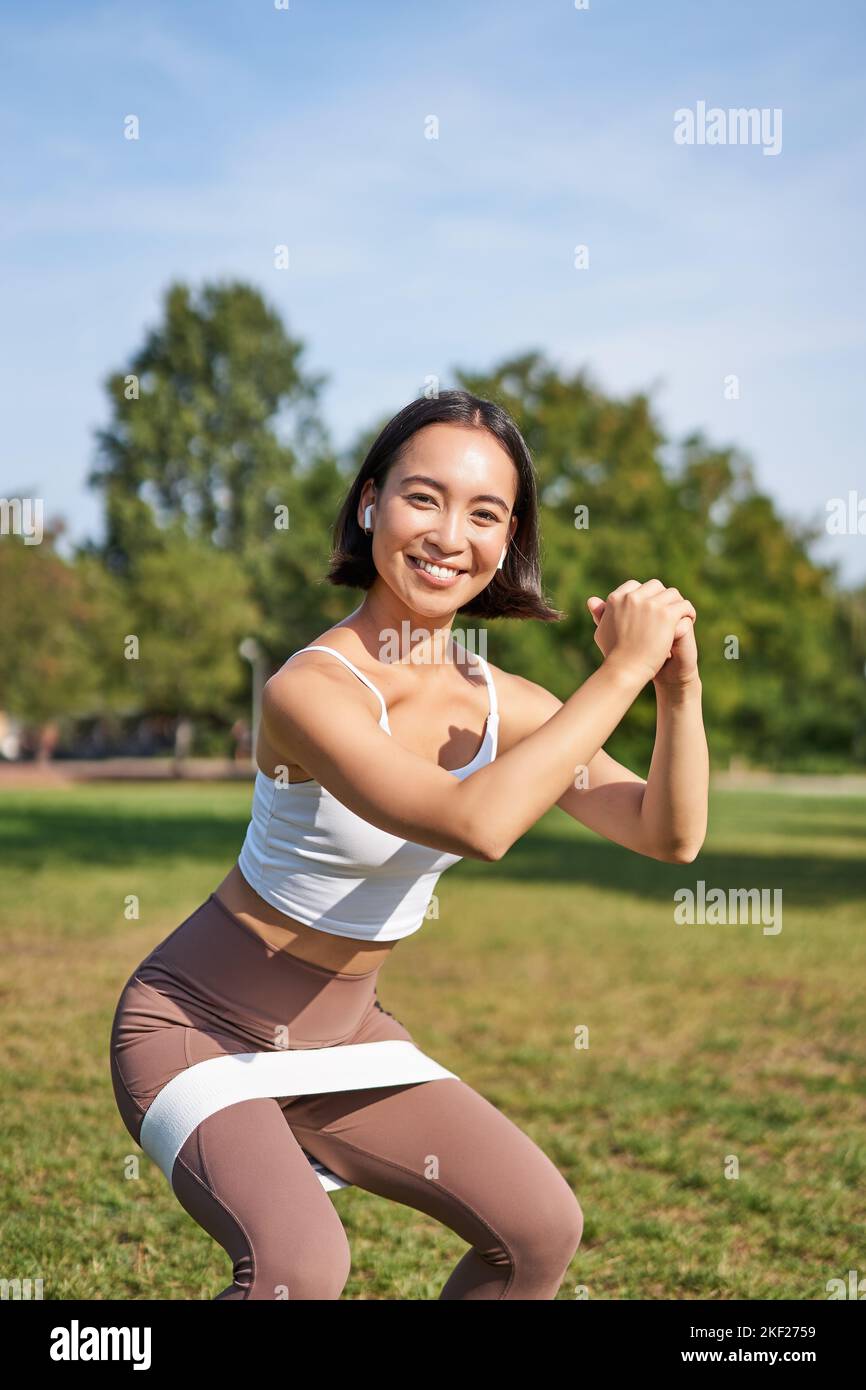 Vertical shot of young fit woman does squats in park, using stretching ...